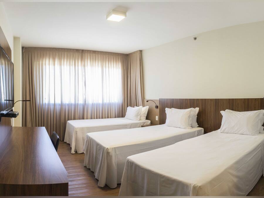 Quarto de hotel com duas camas de solteiro, uma escrivaninha com televisão de tela plana, cabeceira de madeira e uma janela grande com cortinas.