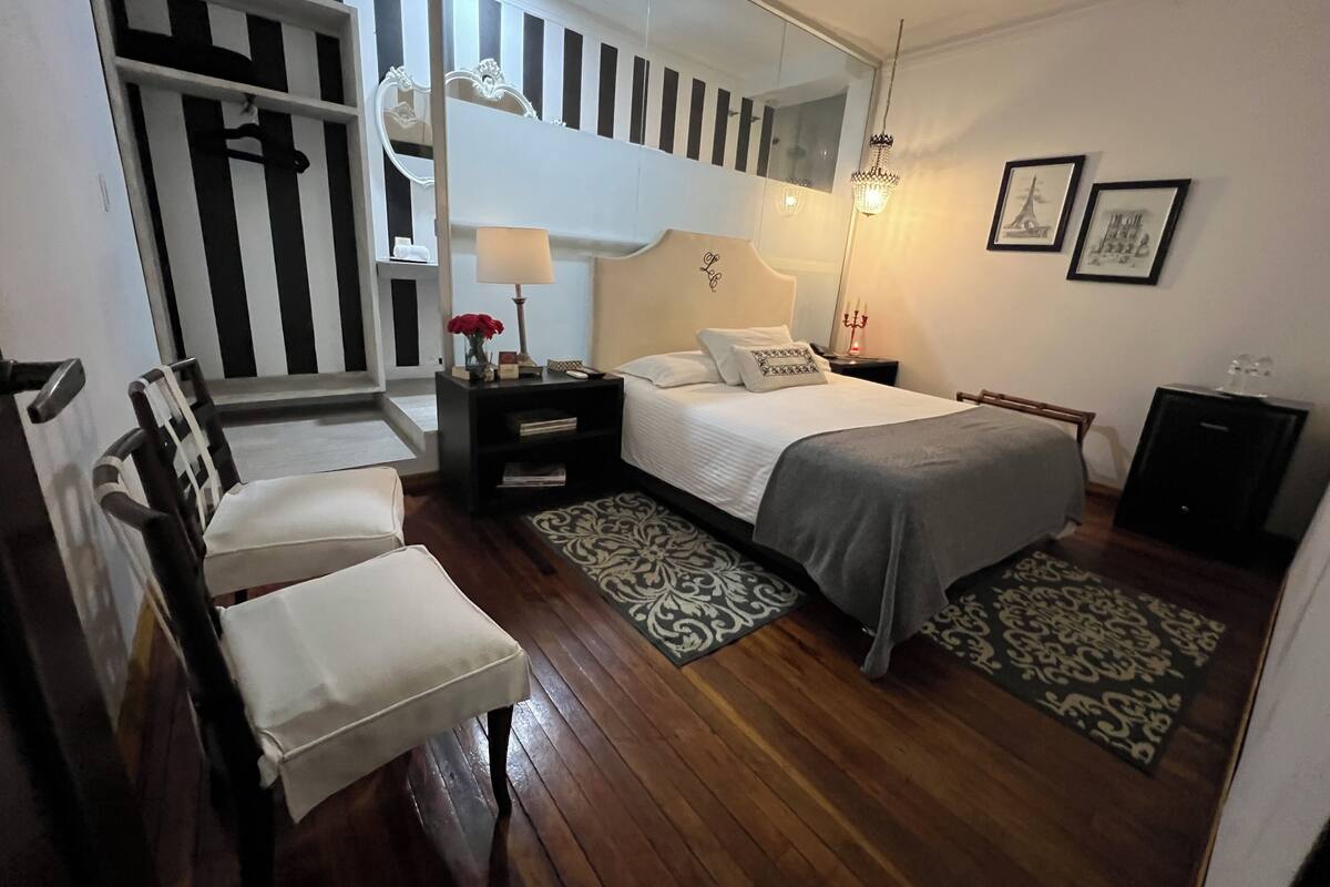 Quarto com cama, mesas de cabeceira, espelho, cadeira e pequena escrivaninha.