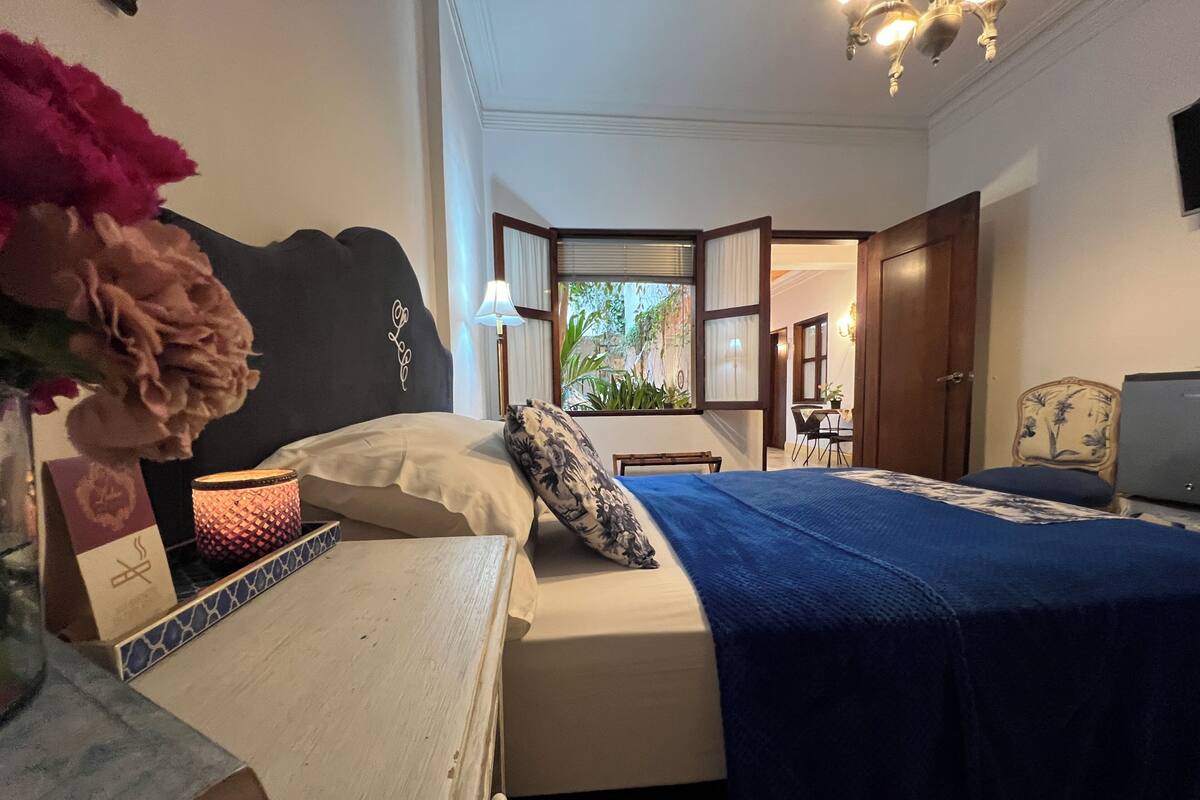 Quarto com cama, mesa de cabeceira, abajur e vista para a sala de jantar.