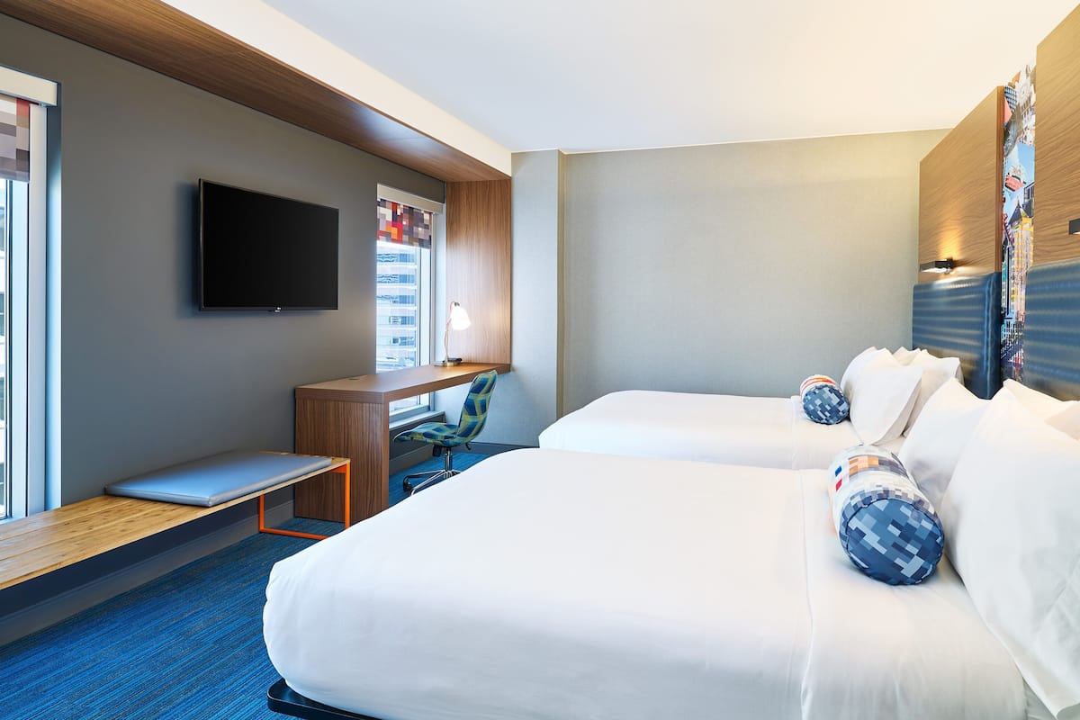 Quarto de hotel com duas camas, uma escrivaninha com um laptop, uma cadeira, uma televisão na parede e uma janela com vista para edifícios.