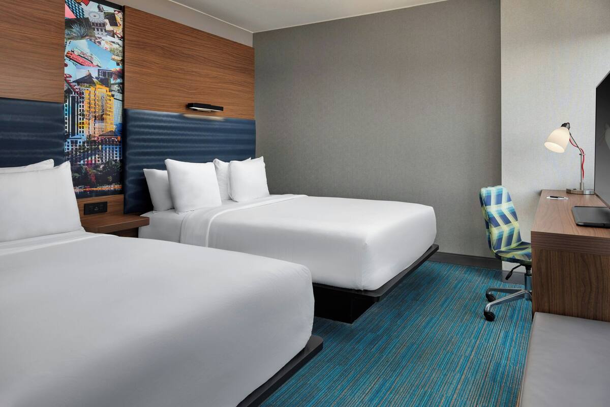 Quarto de hotel com duas camas, uma escrivaninha, uma cadeira, um abajur e uma obra de arte com vista para a cidade na parede.