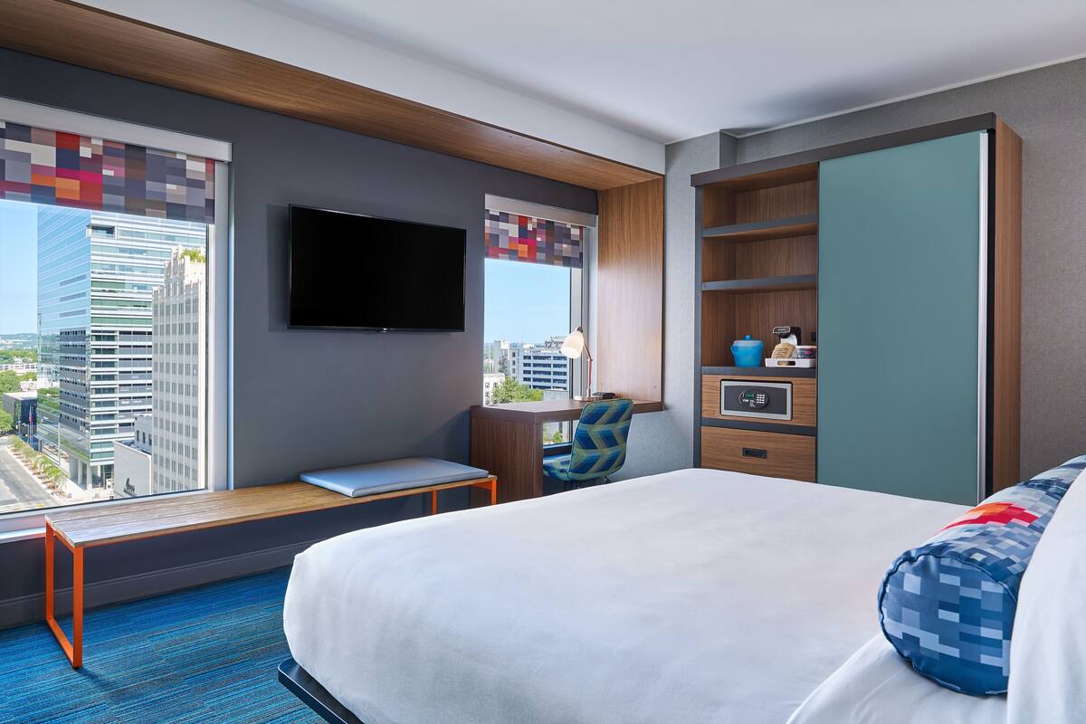 Quarto de hotel moderno com uma cama grande, uma escrivaninha com cadeira, televisão e vista para a paisagem urbana.