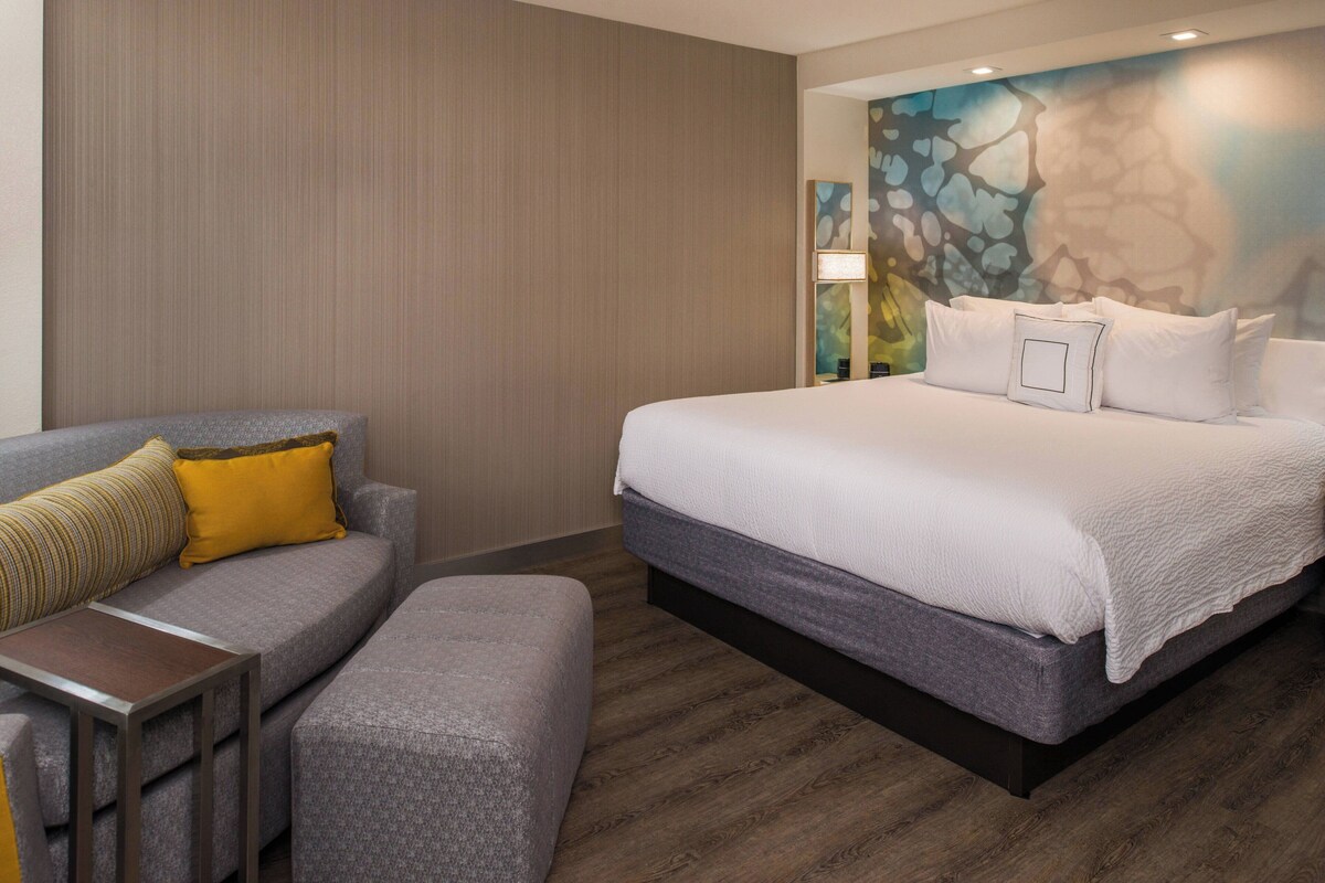 Quarto de hotel moderno com uma cama grande, um sofá cinzento e uma mesinha.