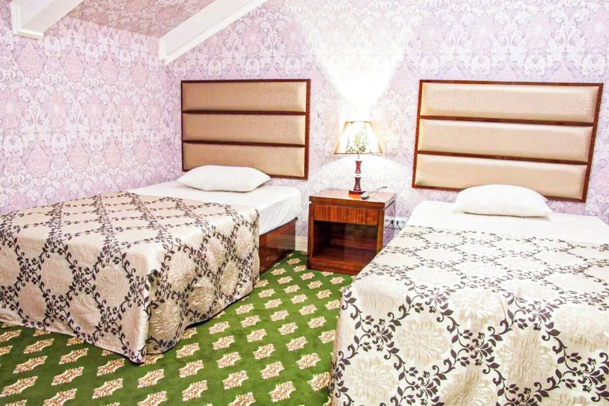 Quarto com duas camas, cada uma com cabeceira e mesa de cabeceira. As paredes têm um papel de parede estampado, e há uma luminária em uma das mesas.