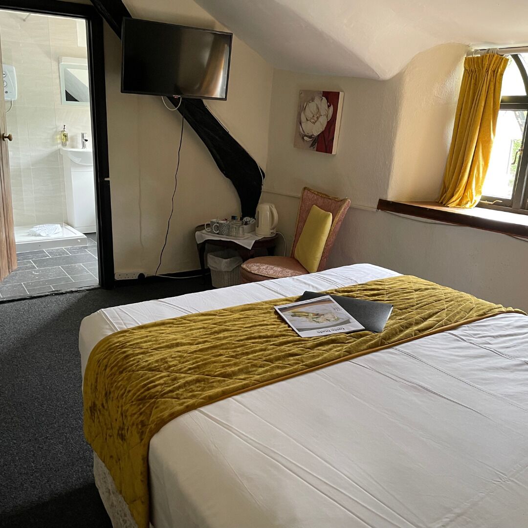 Quarto de hotel com cama, televisão presa à parede, mesinha com chaleira, cadeira e janela com cortinas.