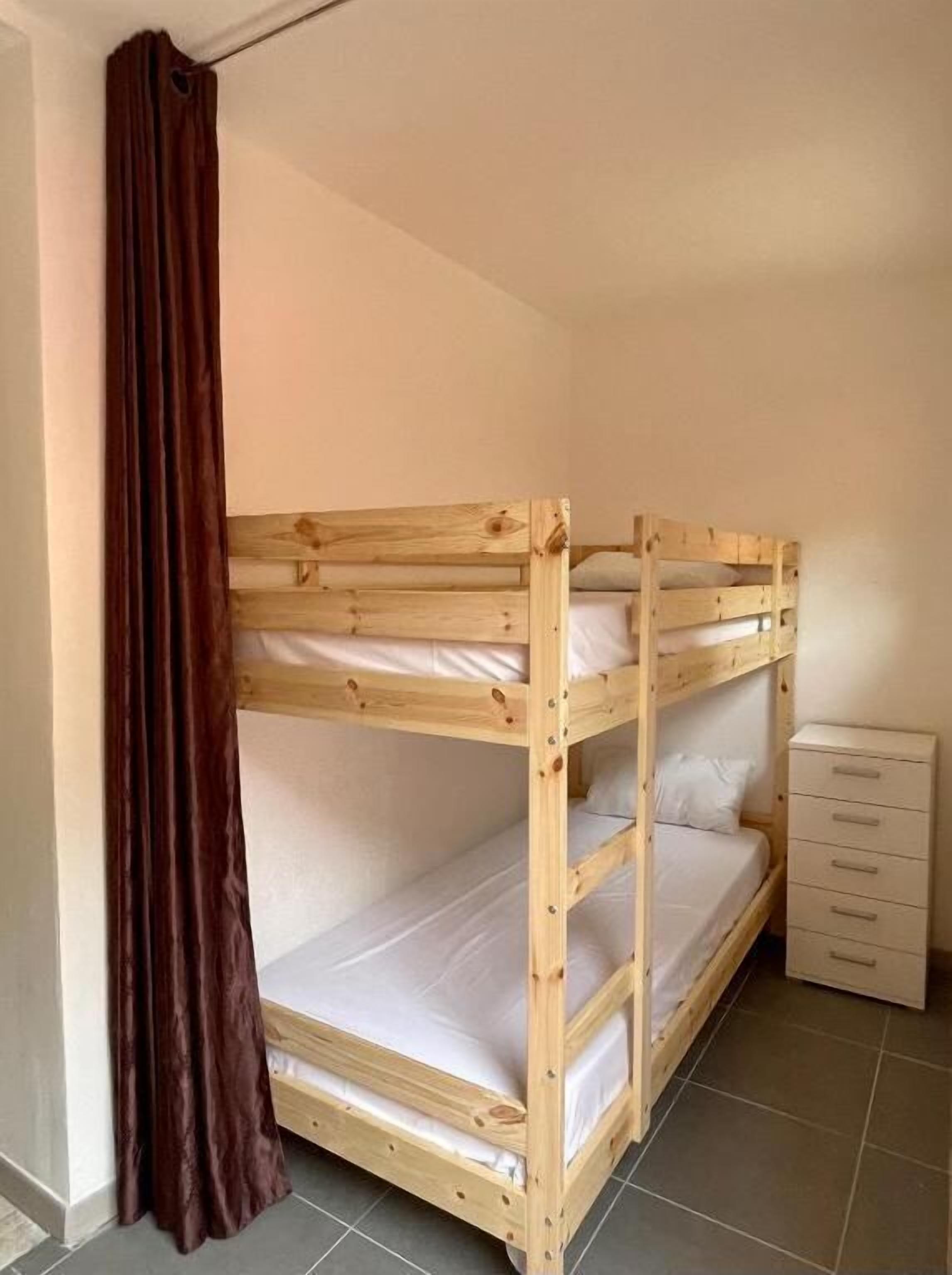 Uma cama beliche com estrutura de madeira, roupa de cama branca e uma cômoda branca com gavetas.