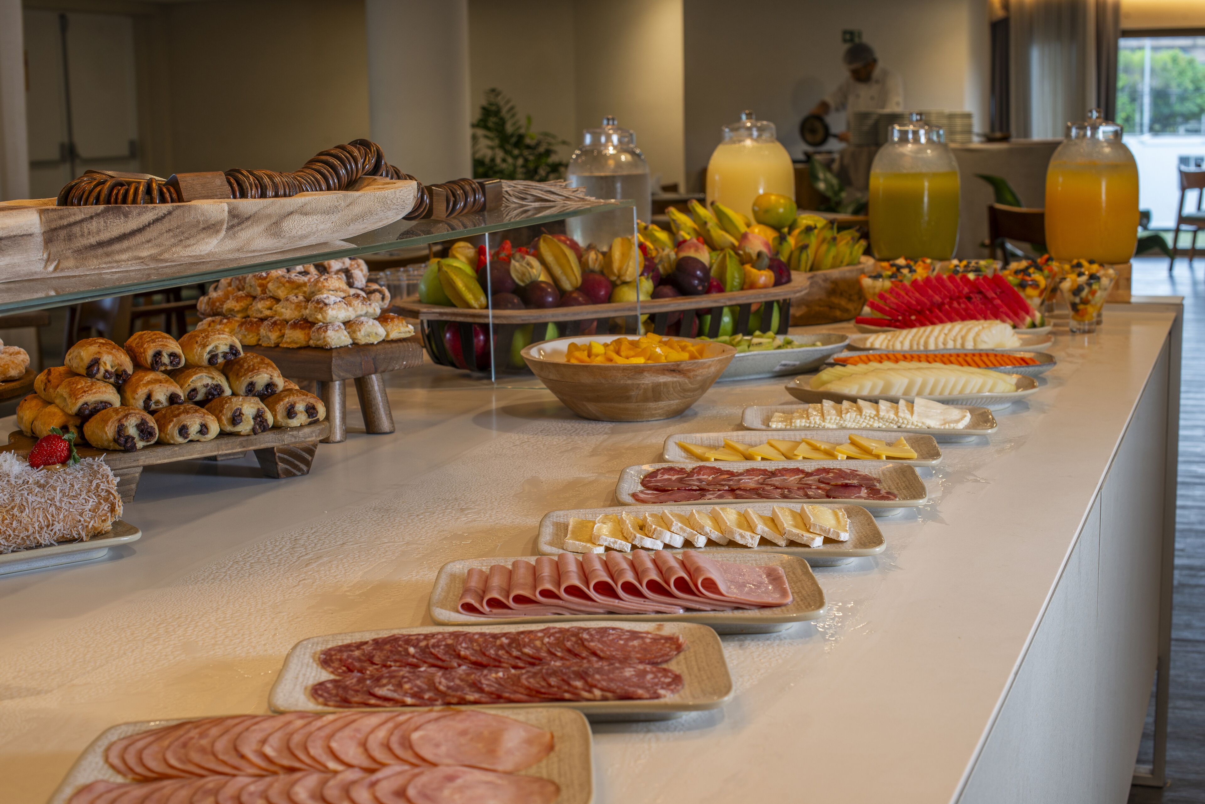 Buffet com diversos itens, incluindo pães, frutas, queijos e carnes.