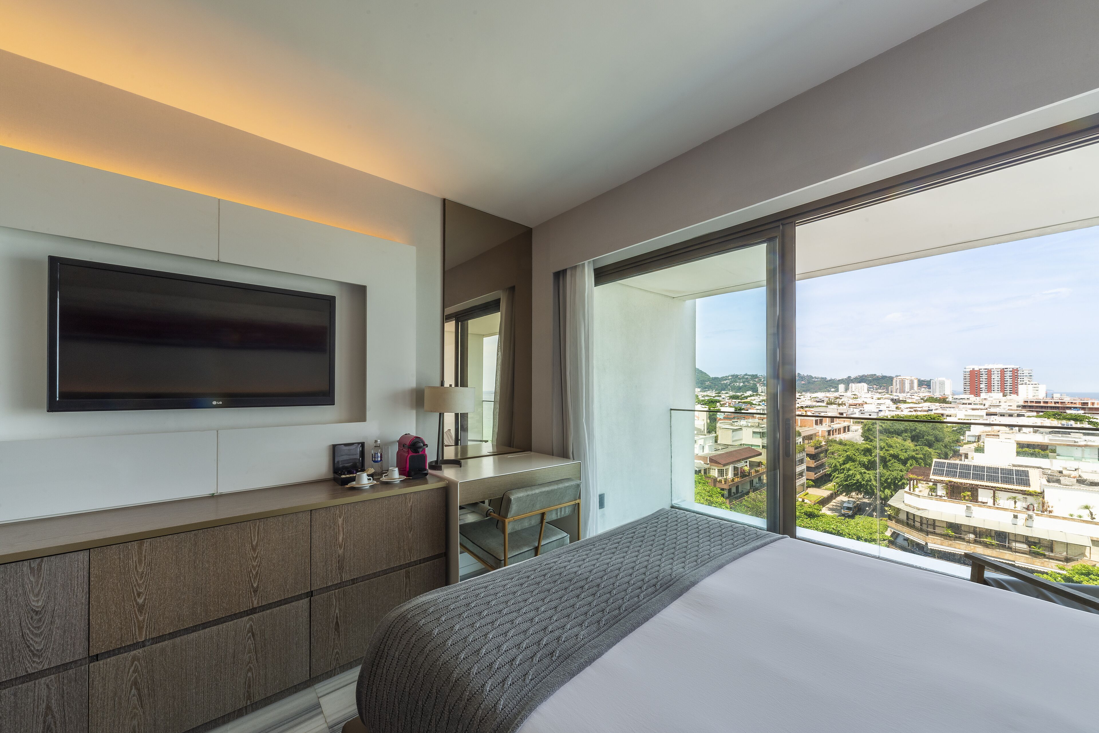 Quarto de hotel moderno com uma cama grande, uma televisão de tela plana na parede, uma escrivaninha com abajur e vista para a cidade através de janelas amplas.