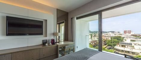 Quarto de hotel moderno com uma cama grande, uma televisão de tela plana na parede, uma escrivaninha com abajur e vista para a cidade através de janelas amplas.