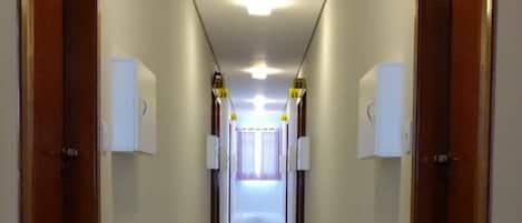 Corredor de hotel com portas numeradas, piso ladrilhado e uma luminária clara.
