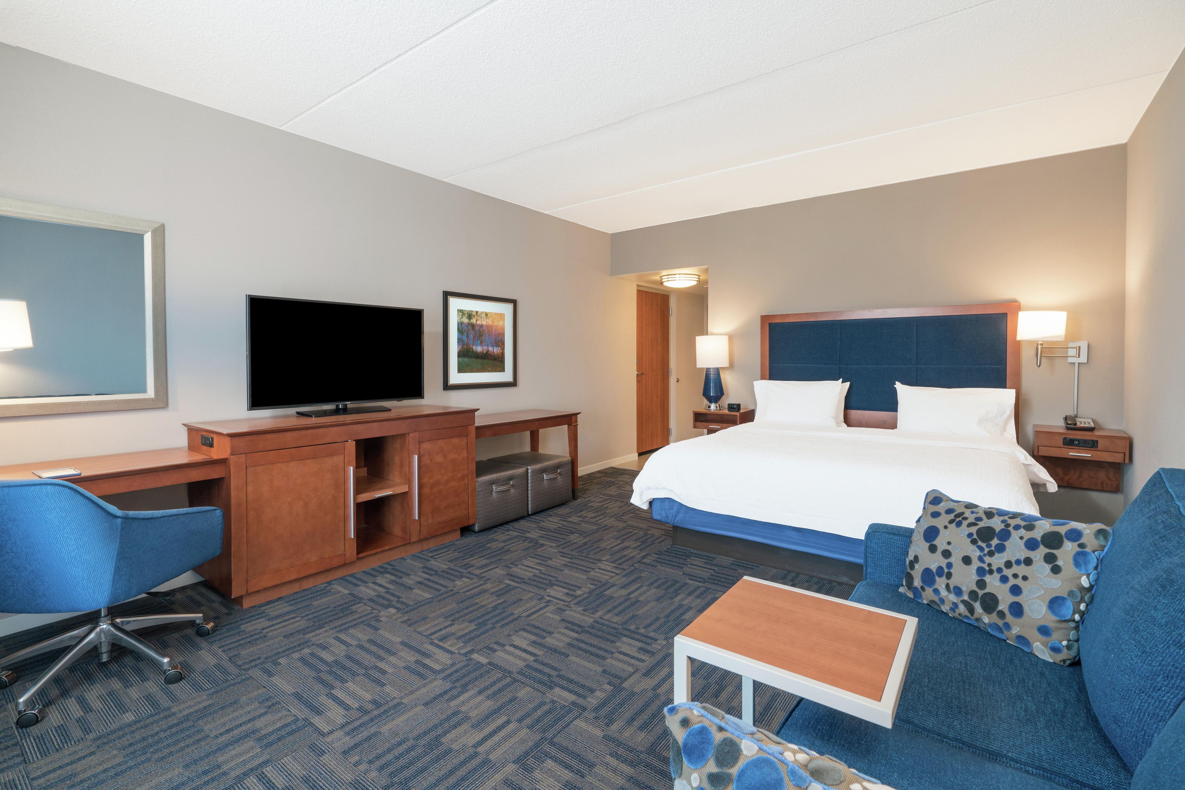 Quarto de hotel com cama, escrivaninha com televisão, cadeira, mesinha e sofá.