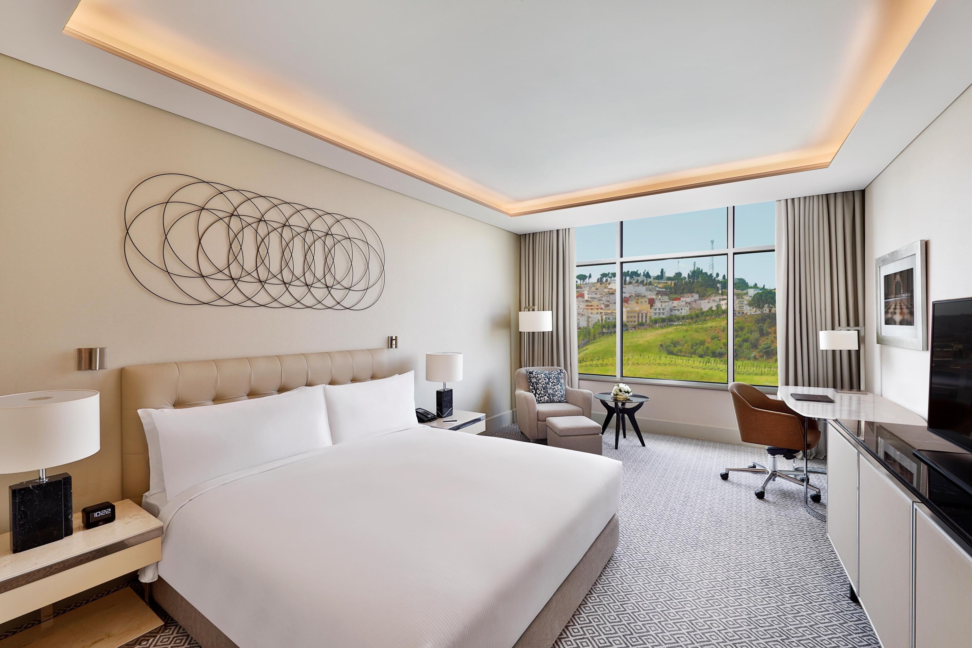 Quarto de hotel com uma cama grande, uma escrivaninha, uma cadeira, uma TV e vista para a cidade.