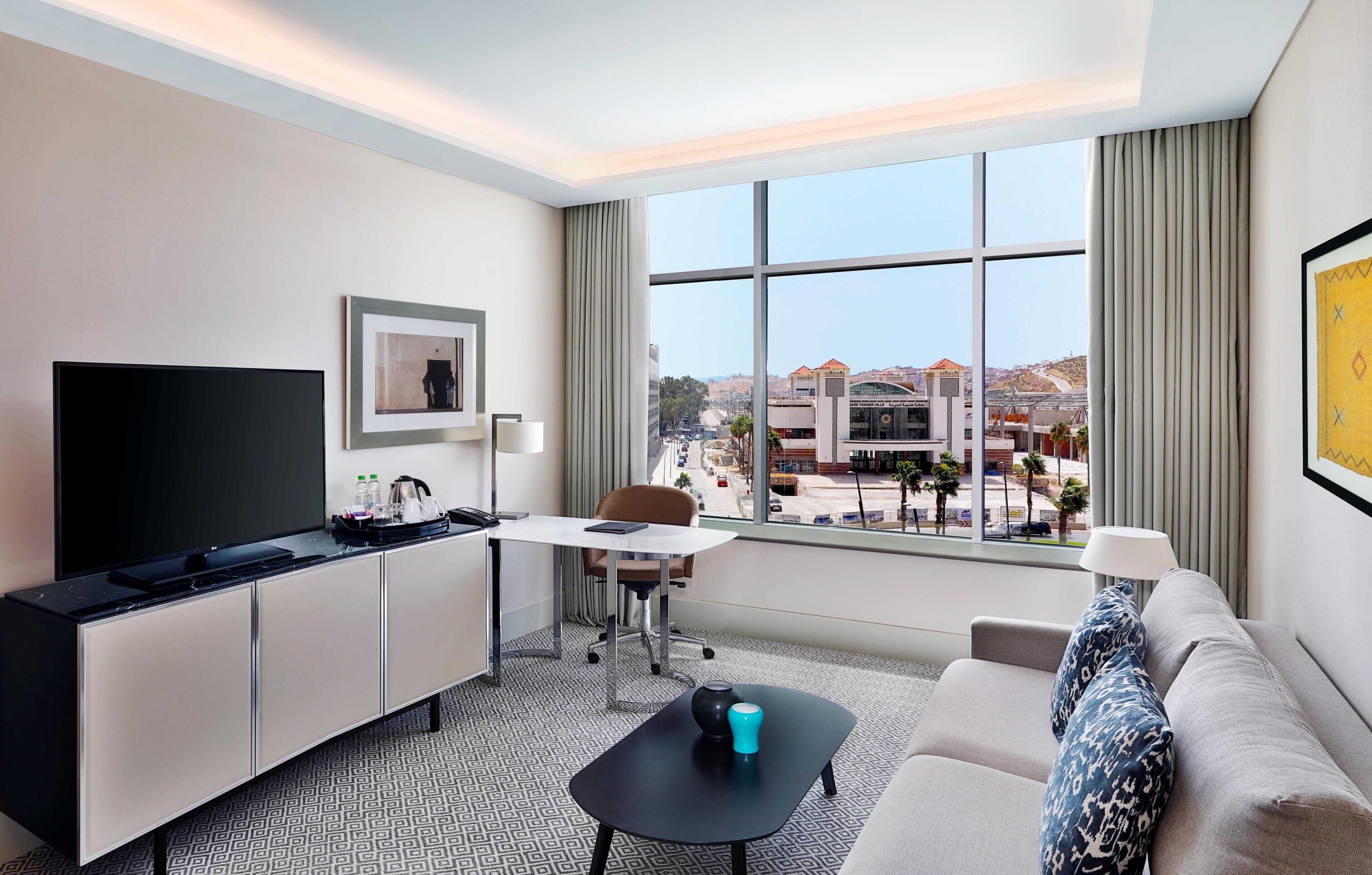 Quarto de hotel com televisão, escrivaninha, cadeira, sofá e grande janela com vista para a cidade.