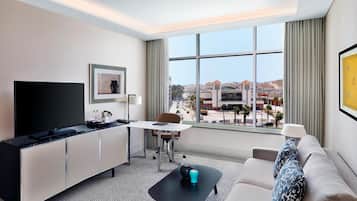 Quarto de hotel com televisão, escrivaninha, cadeira, sofá e grande janela com vista para a cidade.