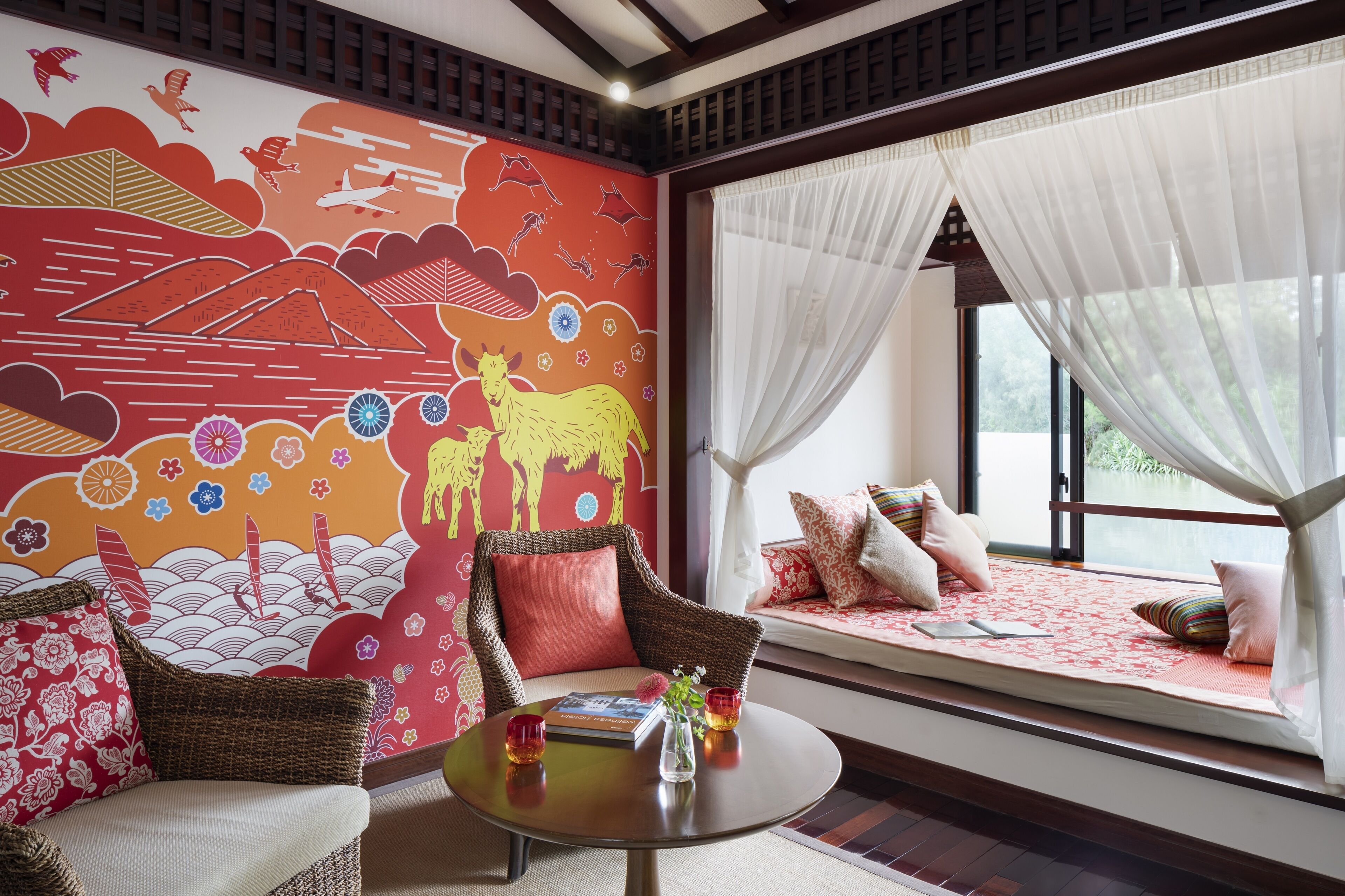 Quarto com um mural grande, um sofá, uma janela com cortinas leves e uma pequena mesa com um livro e um vaso.