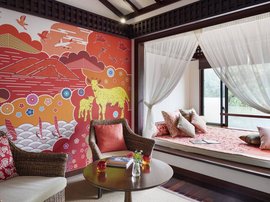 Quarto com um mural grande, um sofá, uma janela com cortinas leves e uma pequena mesa com um livro e um vaso.