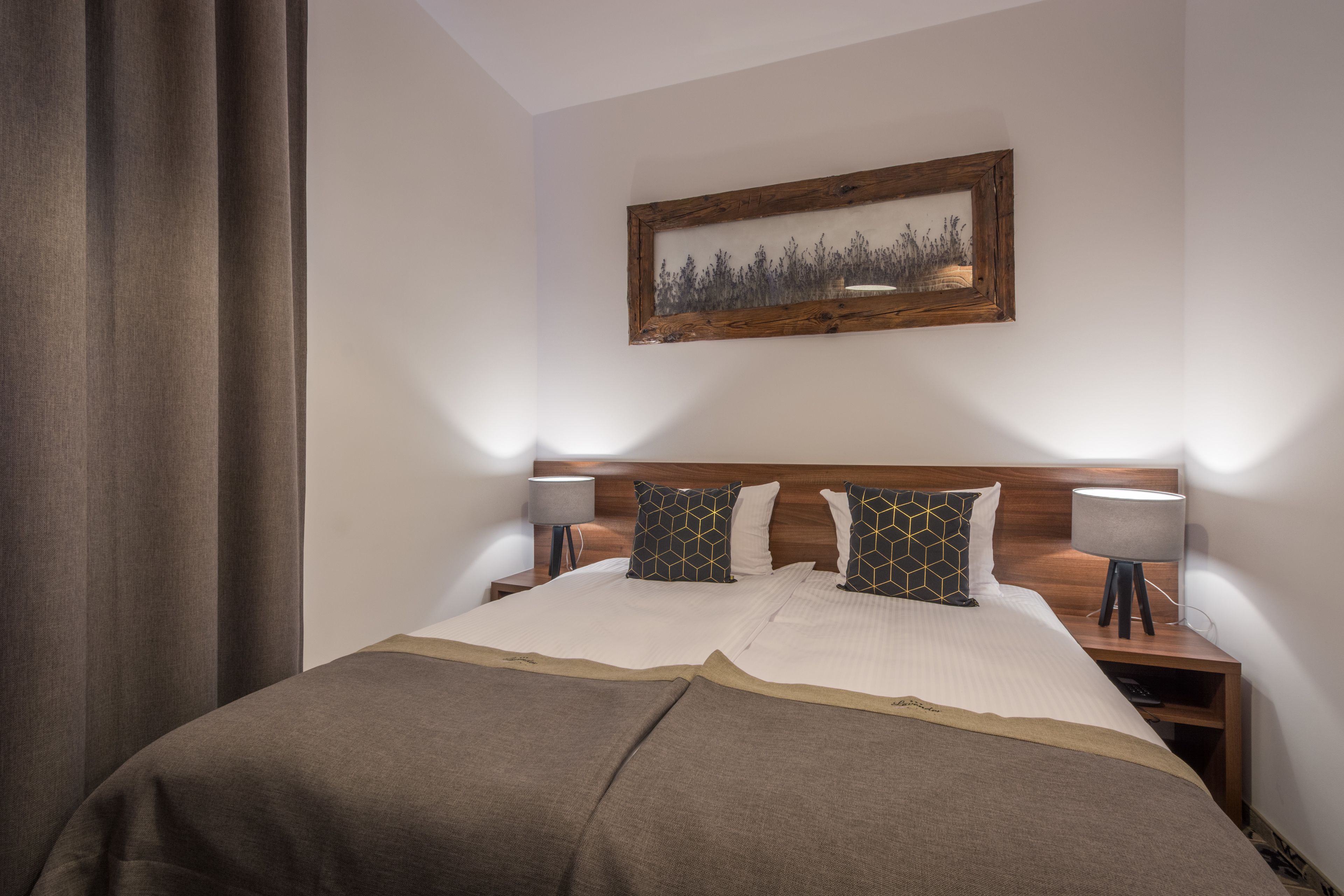 Cama bem arrumada com manta marrom, duas almofadas estampadas e cabeceira de madeira. Há um quadro emoldurado acima da cama, e duas luminárias de cabeceira de cada lado.