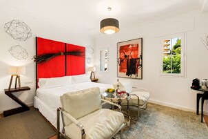 Quarto de hotel moderno com uma cama grande, uma parede de destaque vermelha, uma área de refeições com uma mesa pequena e cadeiras, e obras de arte nas paredes.