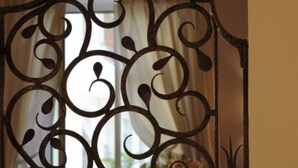 Um portão de ferro forjado com detalhes elaborados, dando acesso a um espaço com vista para uma sala de jantar e uma janela com cortinas.