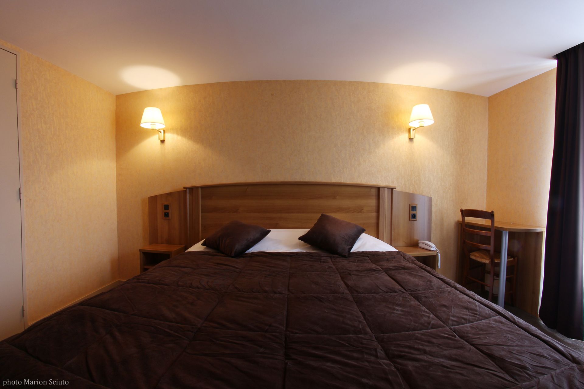 Quarto de hotel com uma cama grande, duas luminárias de cabeceira, um painel de madeira, mesa de cabeceira e uma cadeira.