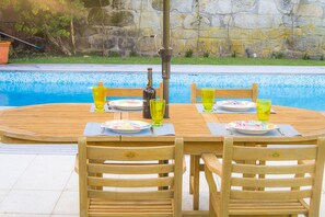 Uma mesa de madeira com cadeiras junto a uma piscina, com pratos, copos e uma garrafa sobre a mesa.
