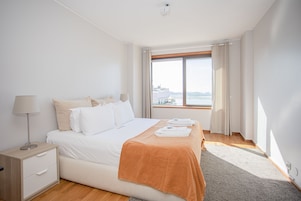 Quarto de hotel moderno com uma cama grande, uma mesa de cabeceira, uma luminária e vista para a cidade através de uma janela ampla.