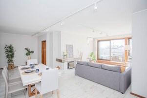 Uma sala de estar moderna com um sofá cinzento, uma mesa de jantar posta com pratos e chávenas, e uma lareira.
