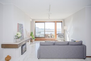 Uma sala de estar moderna com um sofá cinzento, lareira, TV e vista para a cidade através de grandes janelas.
