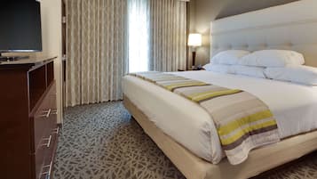 Quarto de hotel com uma cama grande, uma televisão sobre um móvel de madeira, um tapete estampado e uma janela com cortinas.