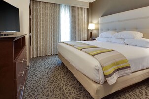 Quarto de hotel com uma cama grande, uma televisão sobre um móvel de madeira, um tapete estampado e uma janela com cortinas.