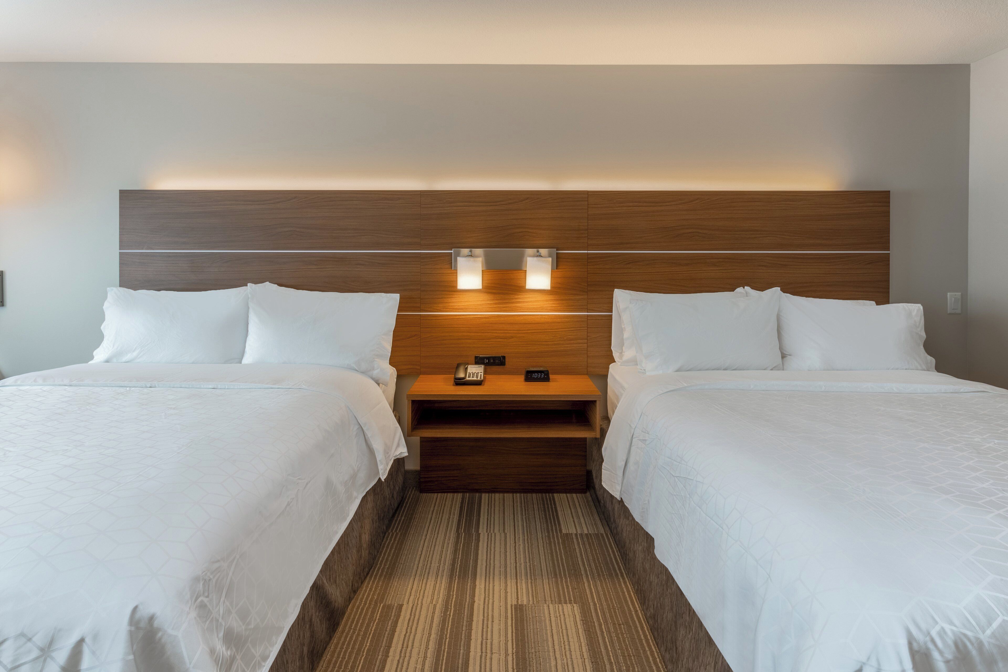 Quarto de hotel com duas camas, cabeceira de madeira, luminárias laterais e mesa de cabeceira com telefone e controle remoto.
