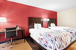 Quarto de hotel com parede vermelha, uma escrivaninha com cadeira e duas camas com roupa de cama branca e colcha estampada.