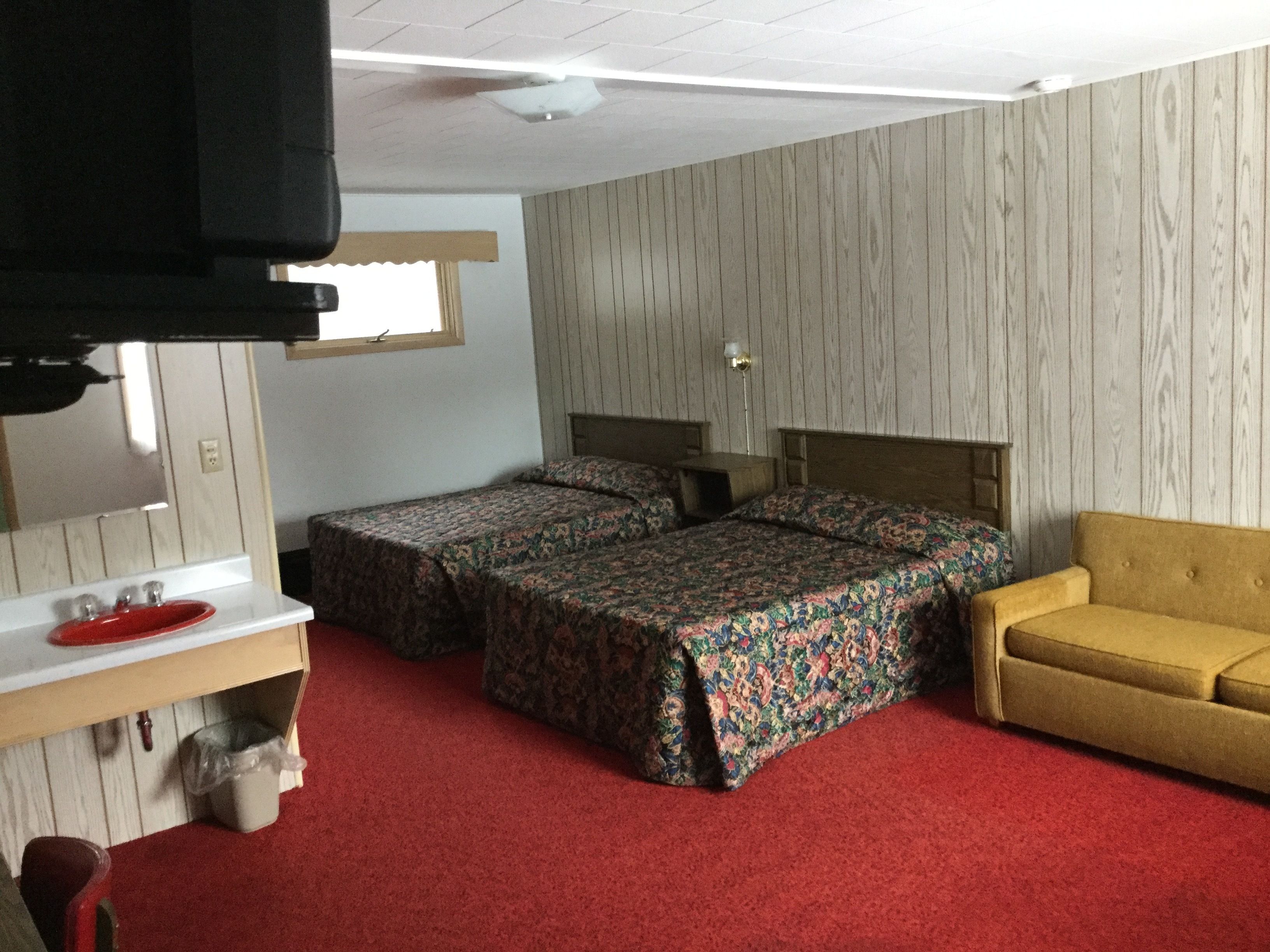 Quarto com duas camas, um tapete vermelho, cabeceira de madeira, sofá amarelo e pia branca.