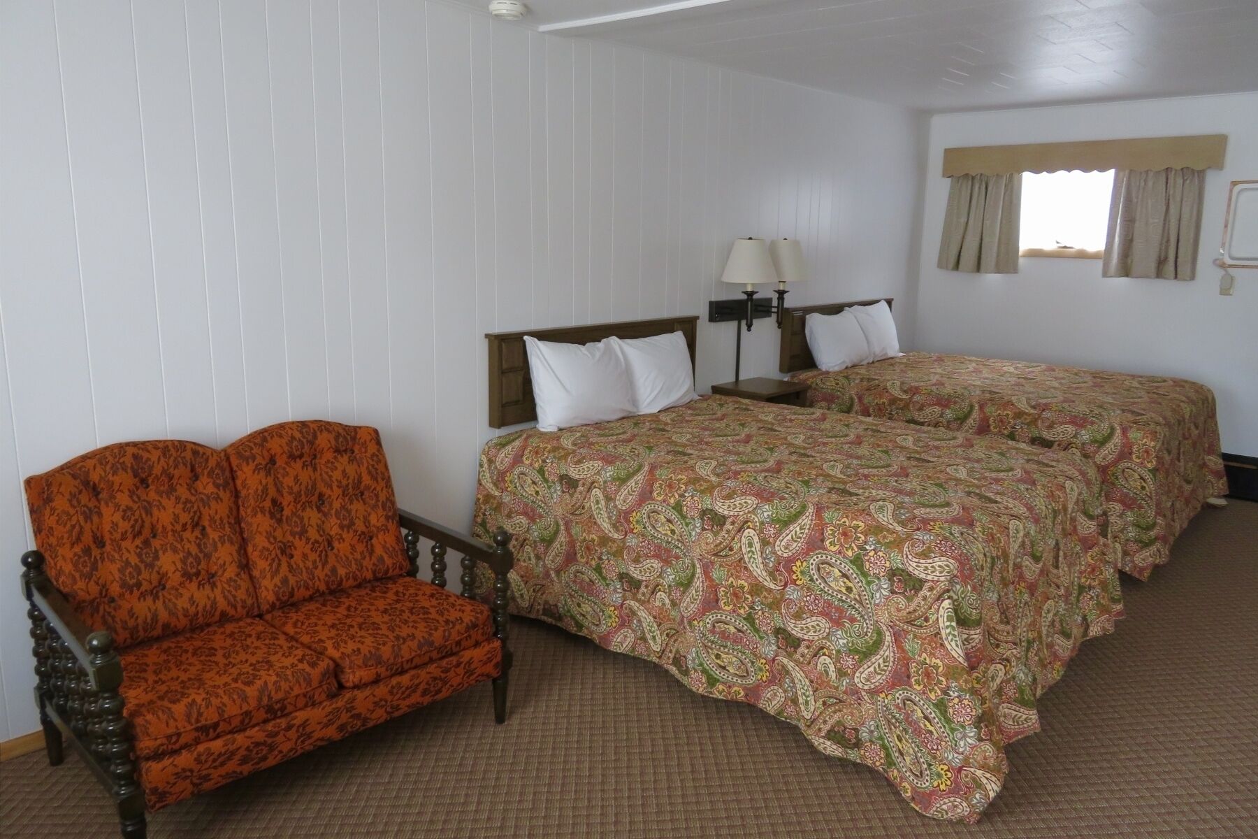 Quarto de hotel com duas camas de solteiro, uma cadeira de madeira e mesa de cabeceira com abajur.