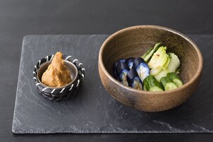 Uma tábua de ardósia com uma tigela de legumes fatiados e uma pequena tigela de um condimento marrom.