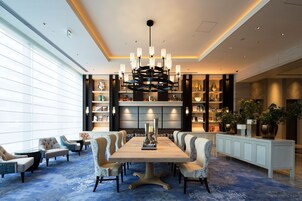 Uma sala de jantar ampla com uma mesa grande rodeada por cadeiras, um lustre moderno e uma estante cheia de livros e objetos decorativos.