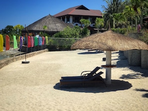 Uma área de praia com guarda-sol de palha, espreguiçadeiras e bandeiras coloridas.