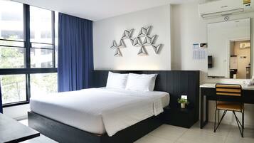 Quarto de hotel moderno com uma cama grande, uma escrivaninha e uma cadeira. Há uma janela com cortinas, ar condicionado fixado na parede e um objeto decorativo na parede.