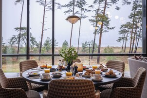 Mesa de jantar posta com itens de café da manhã, cercada por cadeiras estampadas, com vista para árvores altas através de grandes janelas.