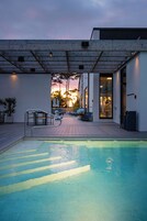 Piscina com degraus de acesso, área de estar coberta com espreguiçadeiras e um edifício com grandes janelas.