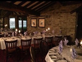 Restaurante com iluminação fraca, paredes de pedra, vigas de madeira e mesas preparadas para refeições.