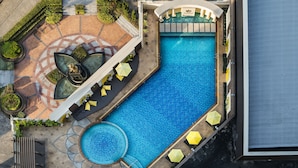 Vista aérea de uma área de piscina de hotel, com uma piscina retangular grande, uma piscina circular menor e uma área de estar com guarda-sóis.