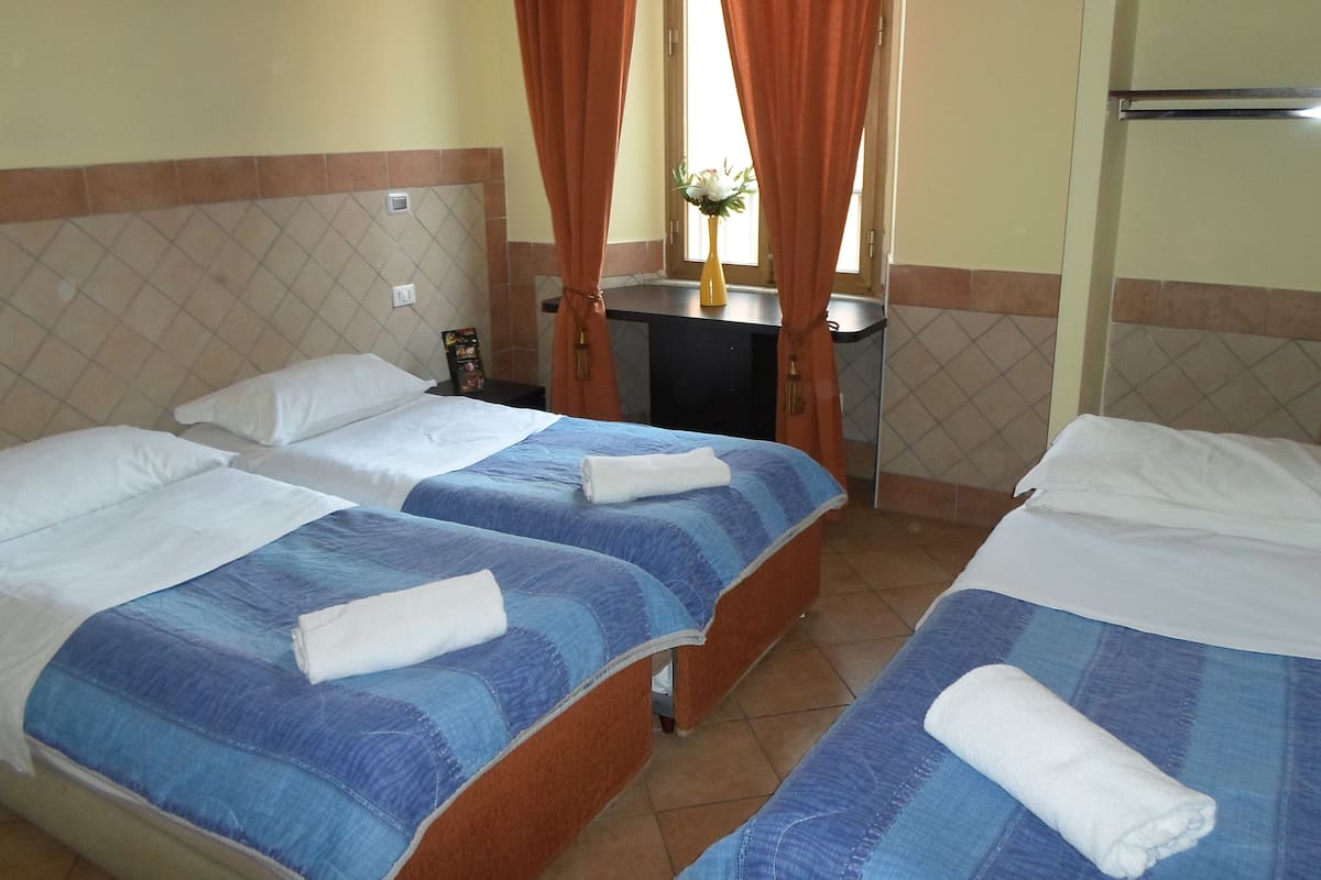 Quarto de hotel com duas camas, cada uma com colcha azul e branca e toalha branca enrolada. Há uma pequena mesa com um vaso de flores e um abajur amarelo.
