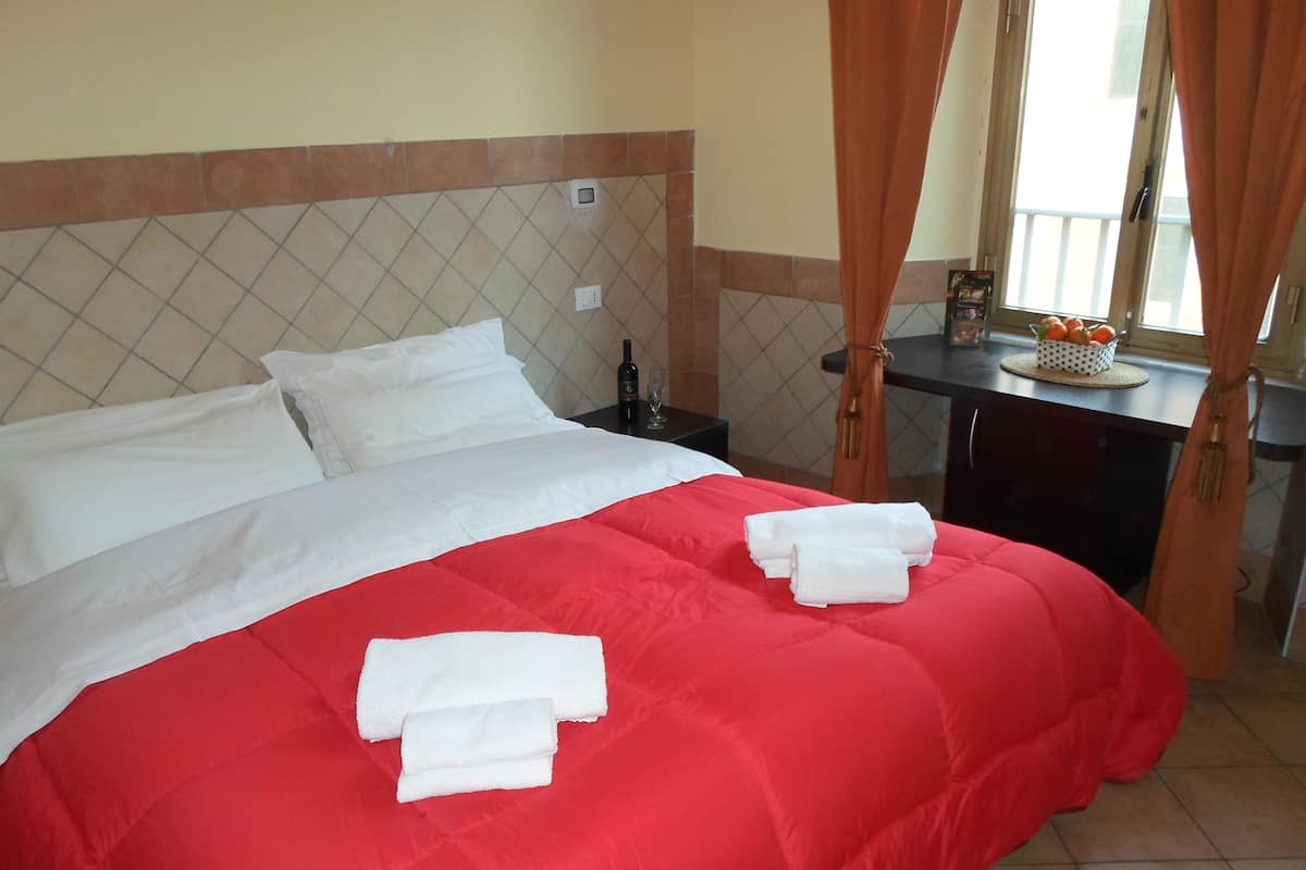 Uma cama bem arrumada com roupa de cama branca e vermelha, uma mesa de cabeceira com garrafa e copos, uma mesinha com frutas e cortinas numa janela.
