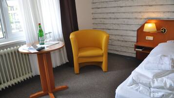 Quarto de hotel com uma cama, uma mesinha redonda com um controle remoto, uma garrafa de água e uma cadeira.