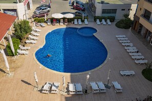 Vista aérea de uma piscina com espreguiçadeiras e guarda-sóis, cercada por um estacionamento e edifícios.