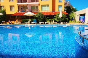 Uma  área de piscina de hotel com espreguiçadeiras e guarda-sóis, rodeada por edifícios com varandas e telhado de telhas vermelhas.