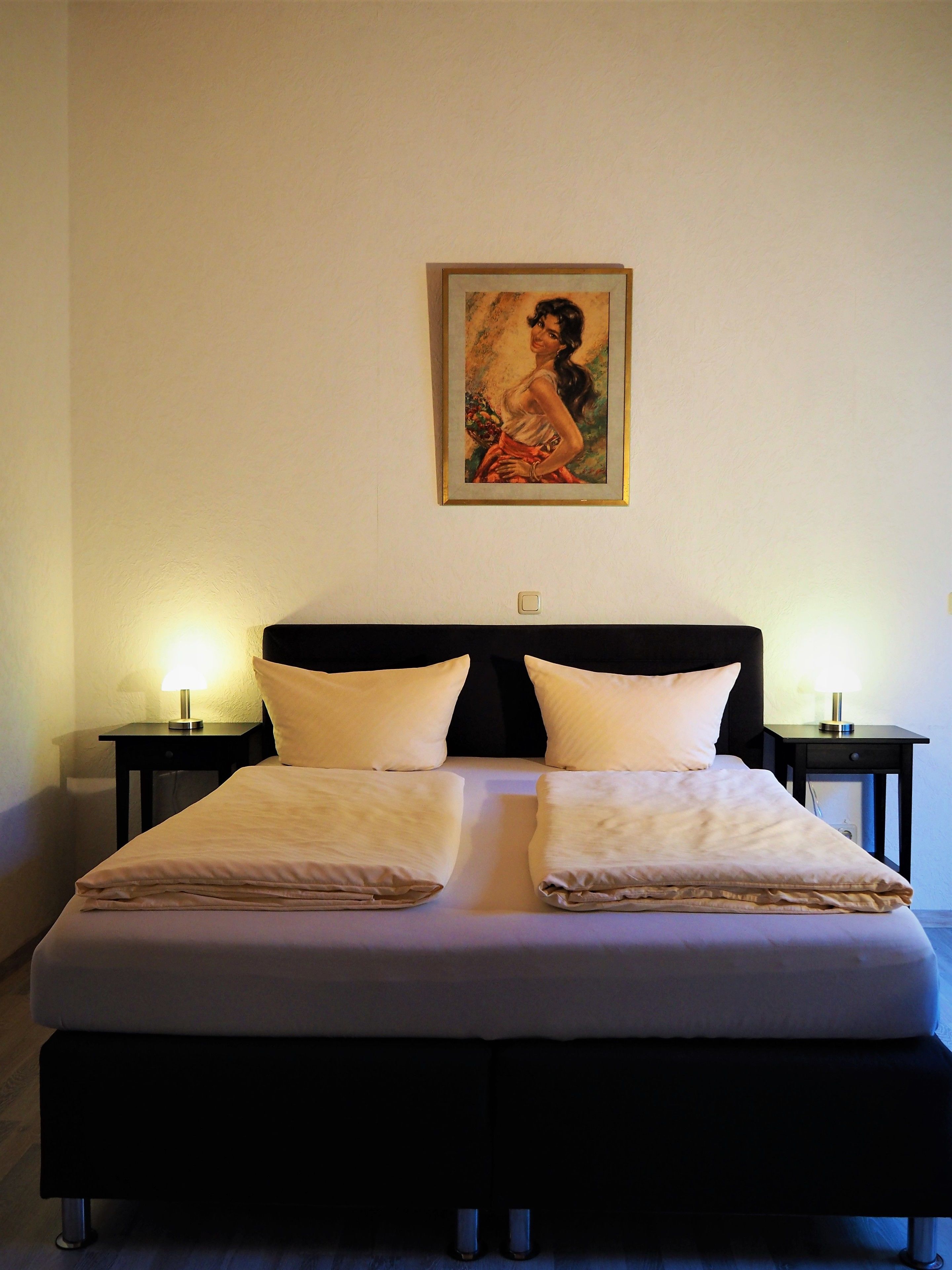 Uma cama de casal com duas almofadas, uma mesa de cabeceira com abajur e um quadro na parede.