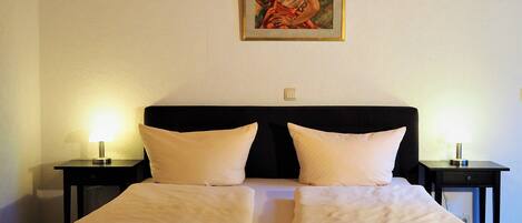 Uma cama de casal com duas almofadas, uma mesa de cabeceira com abajur e um quadro na parede.