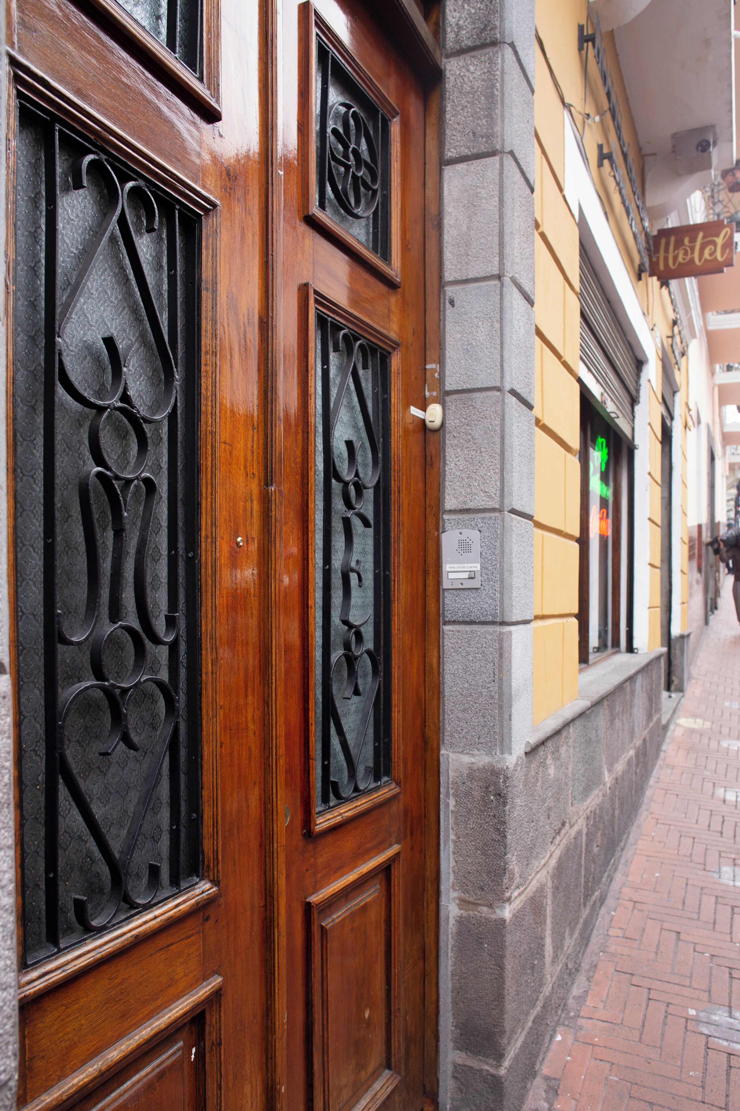 Uma porta de madeira com detalhes metálicos elaborados, uma rua de calçada de tijolos e um edifício com a placa 'Hotel'.
